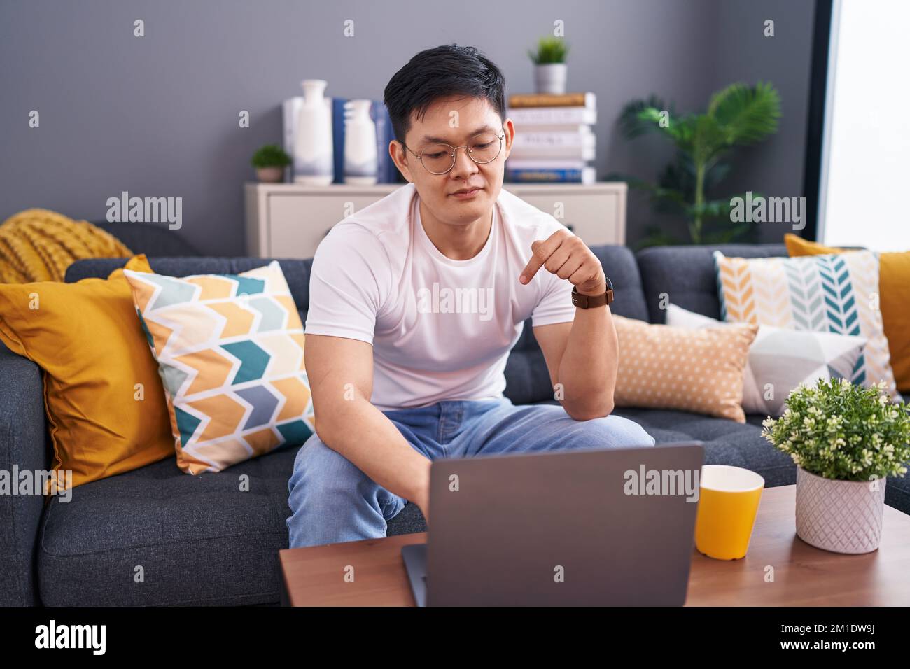 Young asian man using laptop at home sitting on the sofa pointing down ...