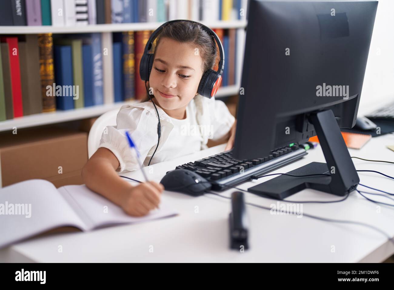 Adorable hispanic girl student using computer and headphones writing on ...
