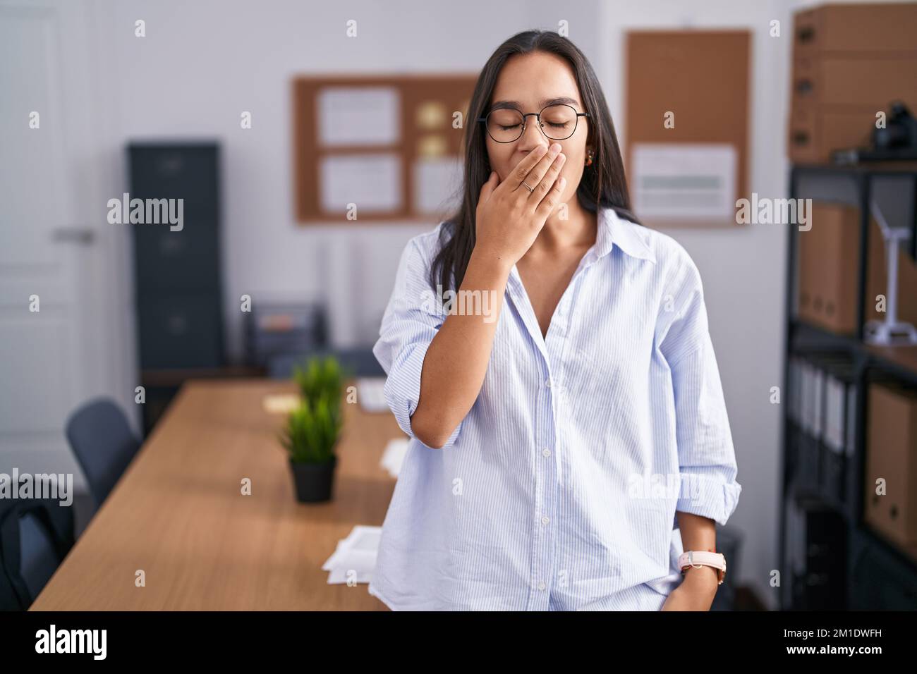 Young hispanic woman at the office bored yawning tired covering mouth ...