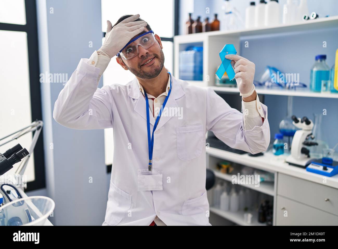 Young hispanic man with beard working at scientist laboratory holding blue ribbon stressed and ...