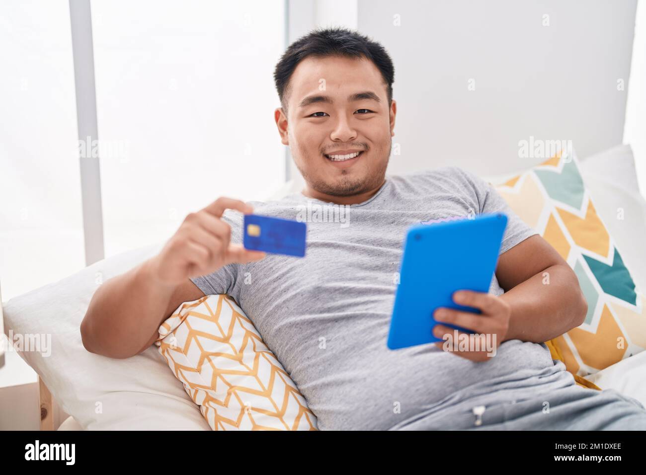 Young chinese man using touchpad and credit card sitting on bed at ...