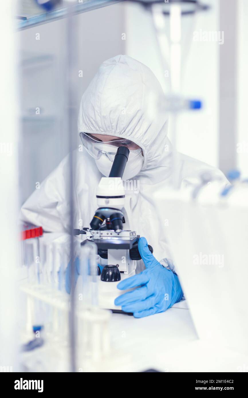 Scientist in sterile ppe suit checking sample of virus under microscope. Chemist in coverall ...
