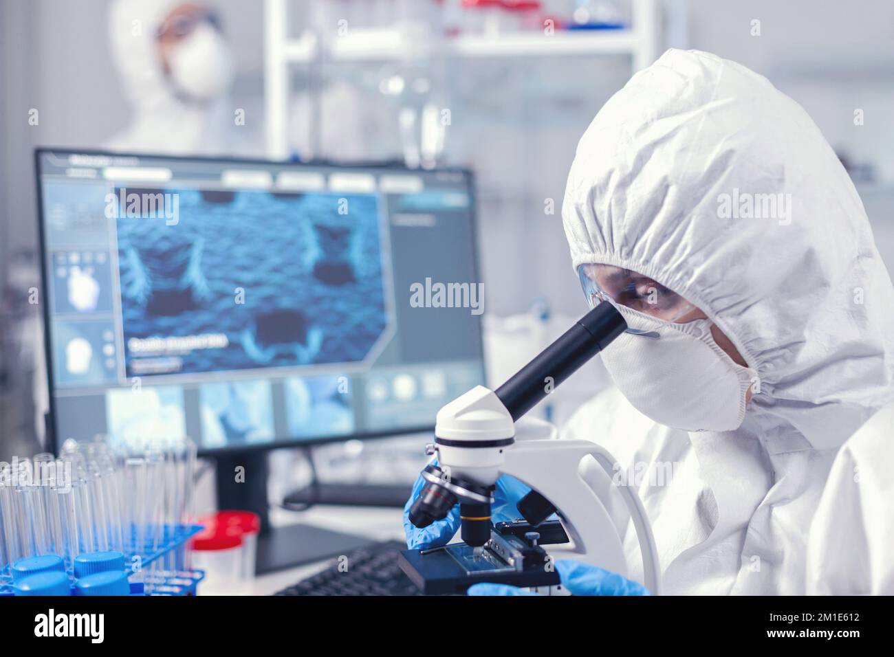 Woman in coverwall sitting at workplace in laboratory using microscope ...
