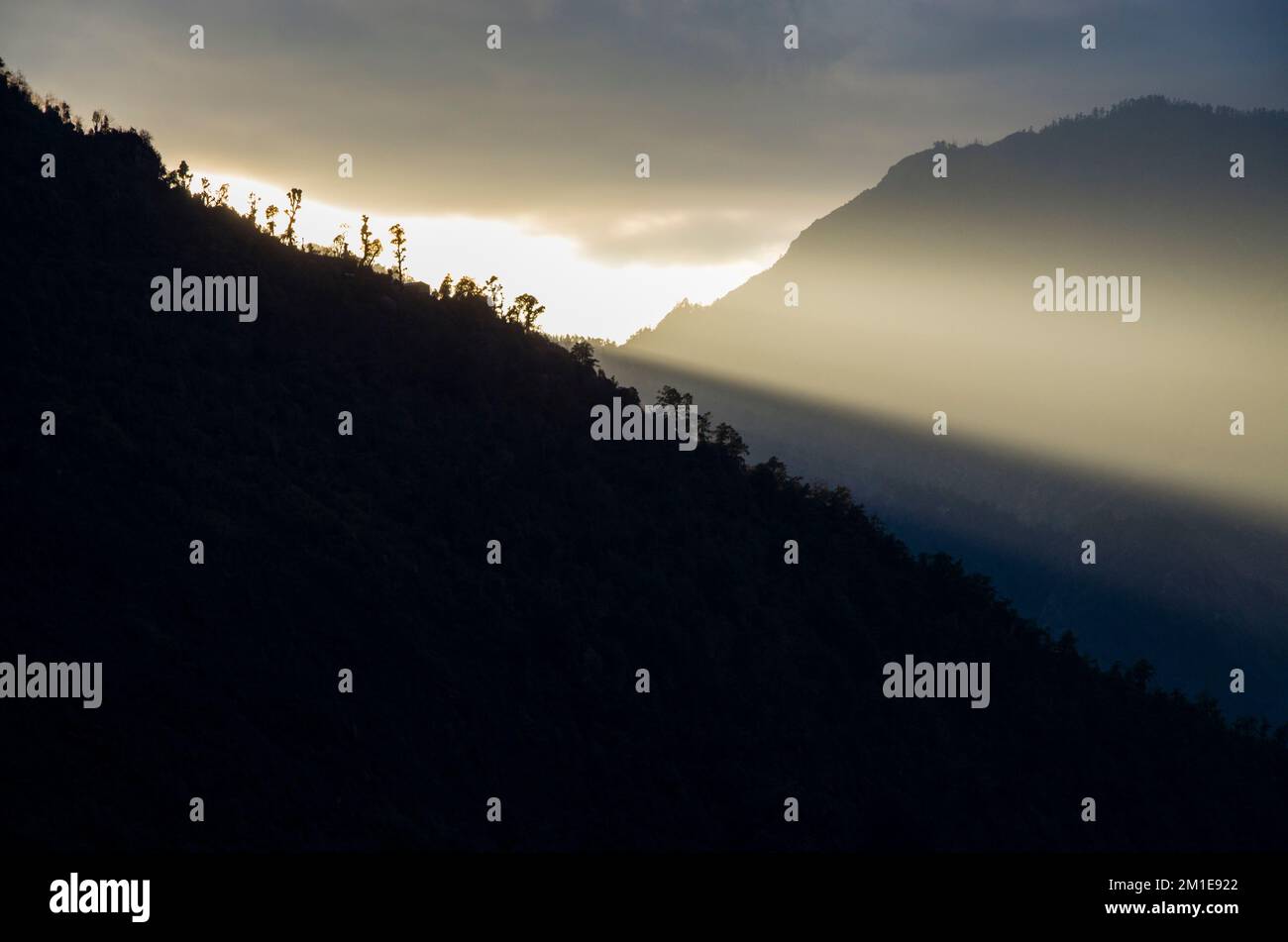 Setting sun beaming through dramatic clouds between 2 mountains Stock ...