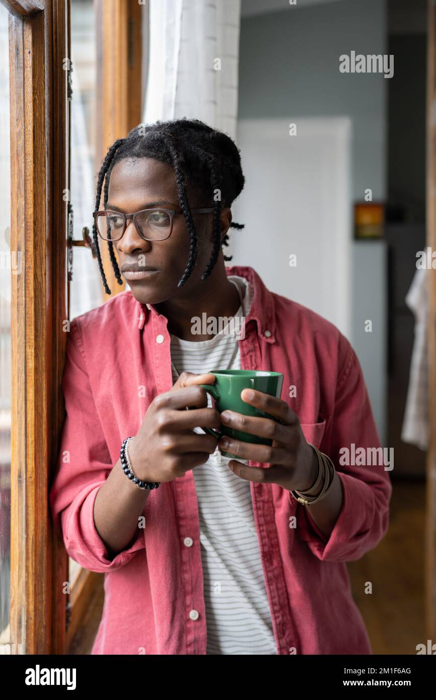 Depressed sad African guy holding cup of tea looking out window, having ...