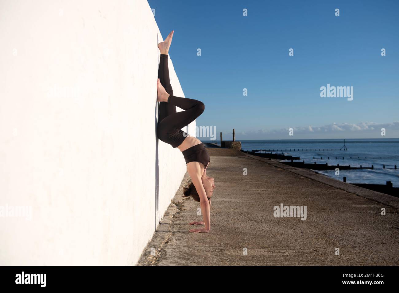 Sporty woman doing a handstand against a white wall by the sea. Outdoor ...