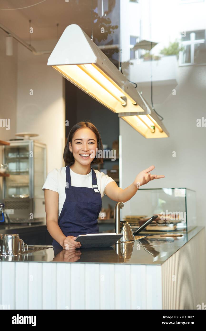 Happy asian girl barista works in coffee shop, waves at client, processing orders behind counter ...