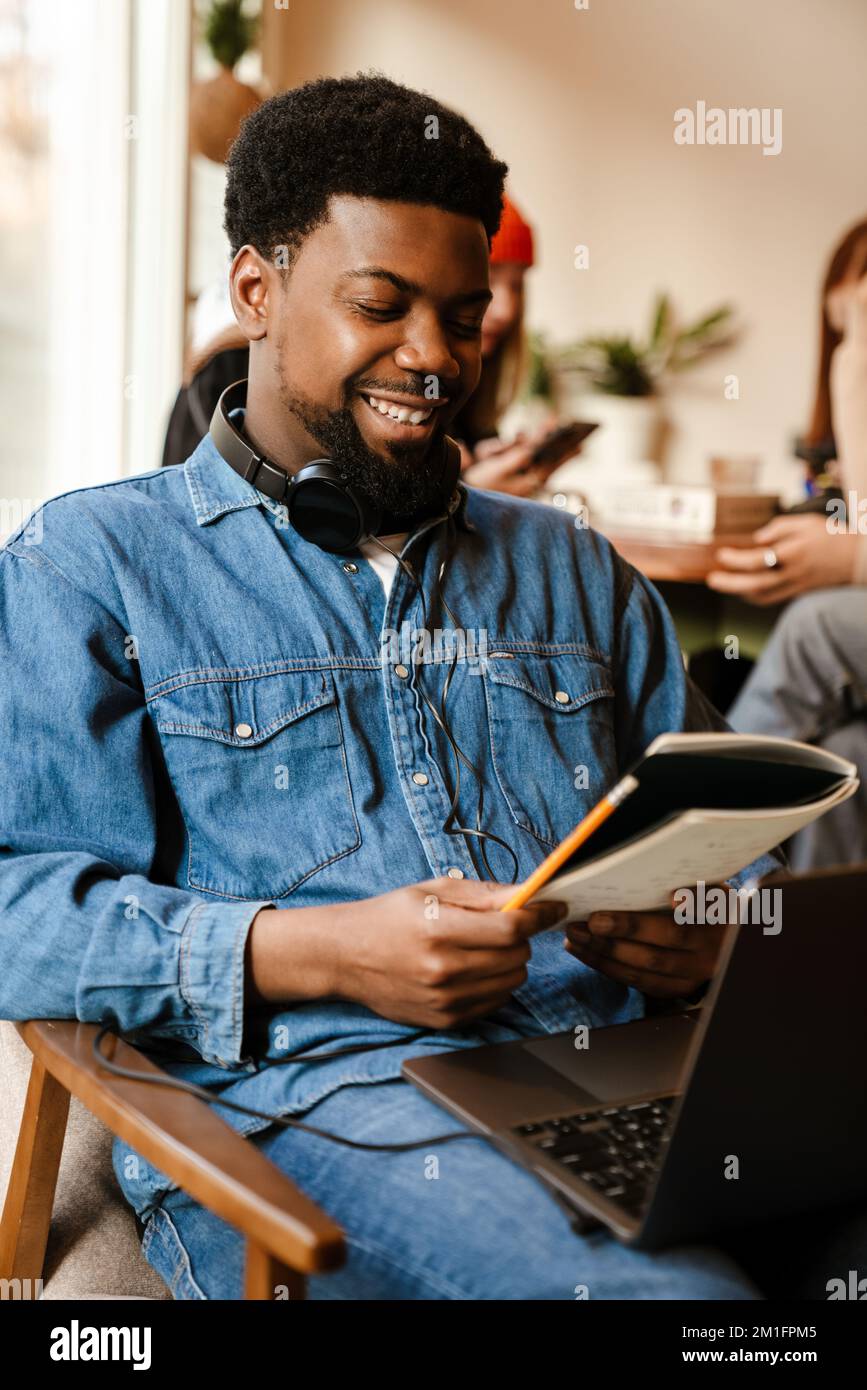 Black bearded man writing down notes while using laptop in cafe indoors ...