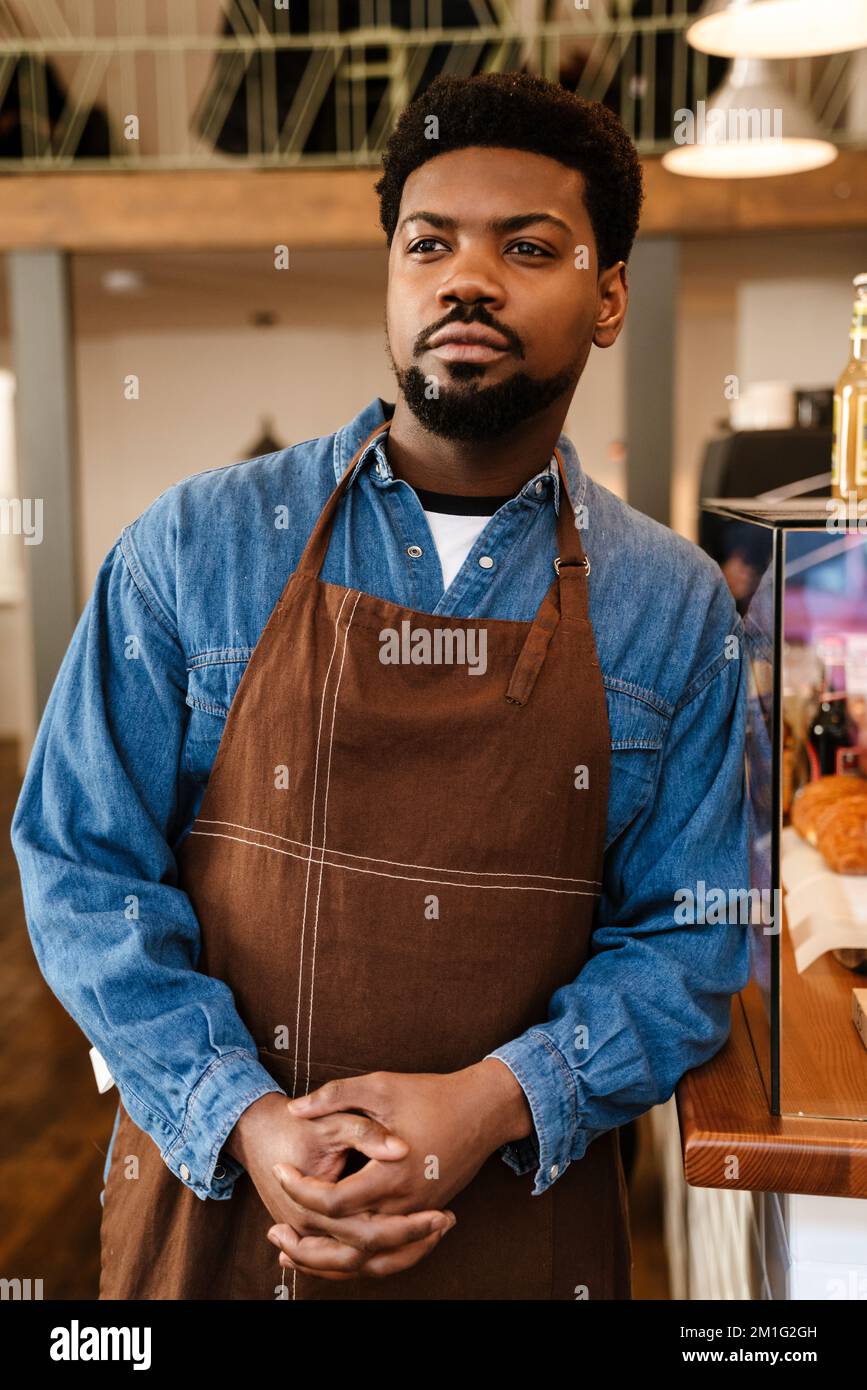 Black bearded man wearing apron standing while working in cafe indoors ...