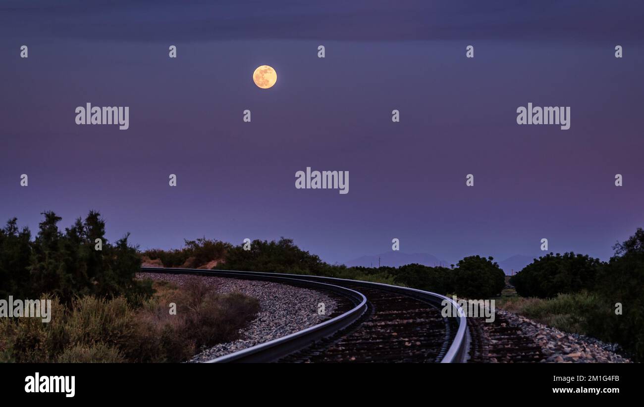 The full moon in purplish blue sunrise sky over winding train tracks in ...