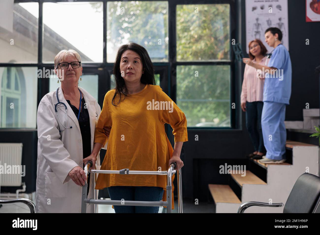 Elderly physician giving support to injured patient trying to walk around clinic with walking ...