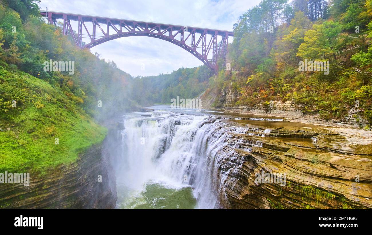 Waterfall cutting through cliffs from drone with steel maroon bridge ...