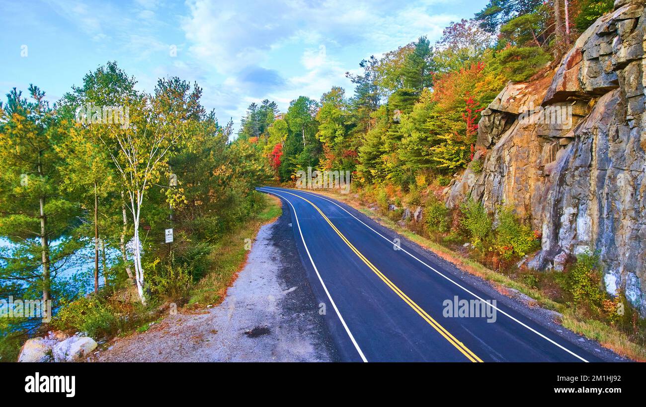 Drone above clean road going through fall forest with lake and cliffs ...