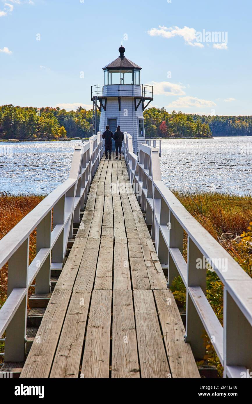 Wood bridge leads to small white Maine lighthouse with two tourists ...