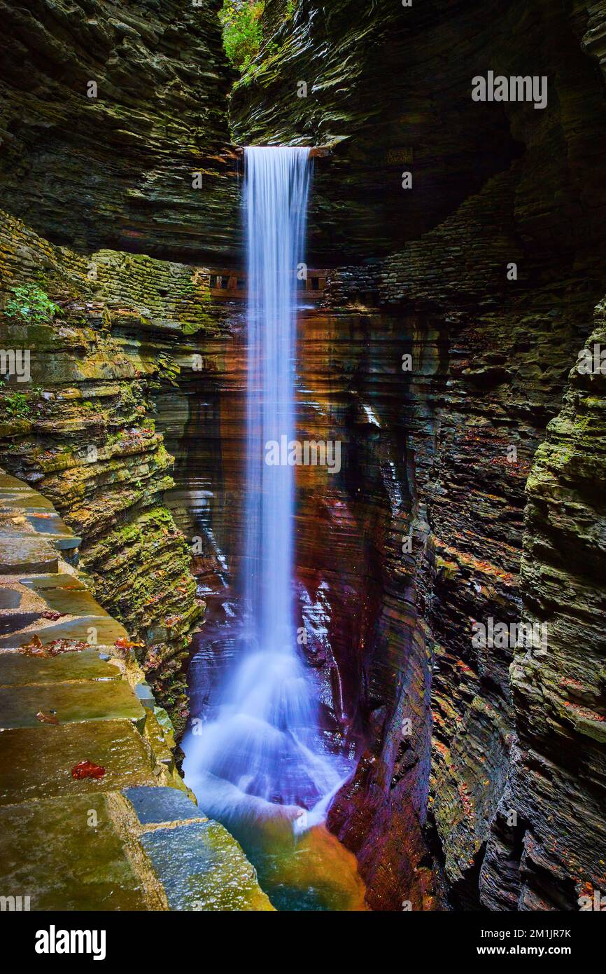 Tall waterfall pours into deep gorge of layered rocks and path leading ...