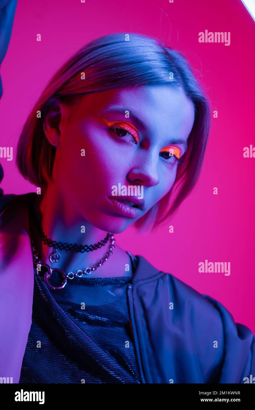 stylish woman in necklaces and glowing neon makeup posing in blue light ...