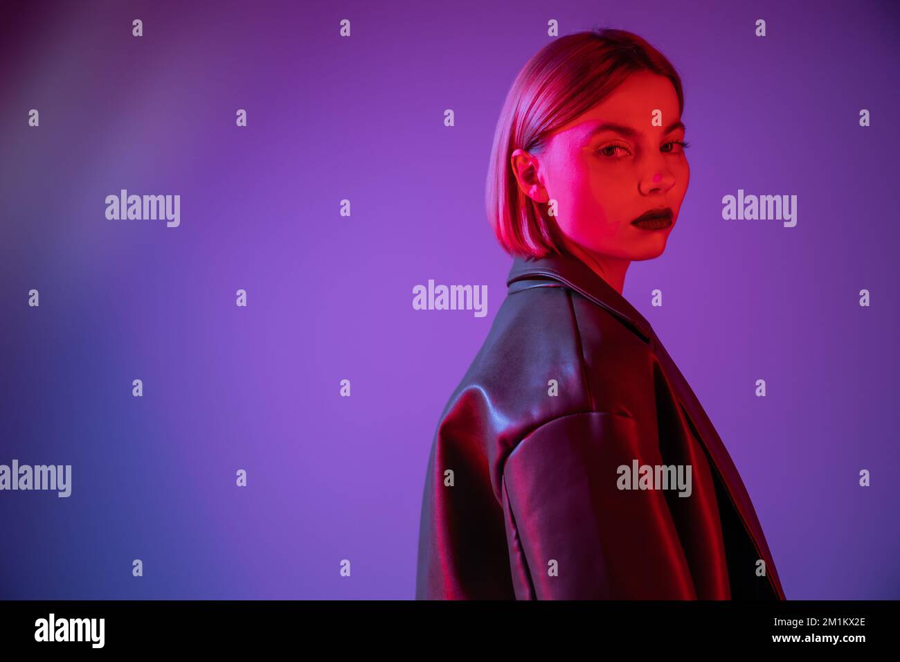 young and stylish woman looking at camera in red light on purple and ...