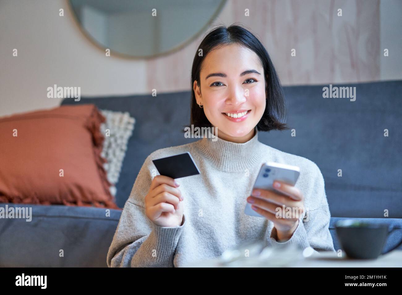 Portrait of smiling korean woman using credit card and smartphone app, paying for purchase ...