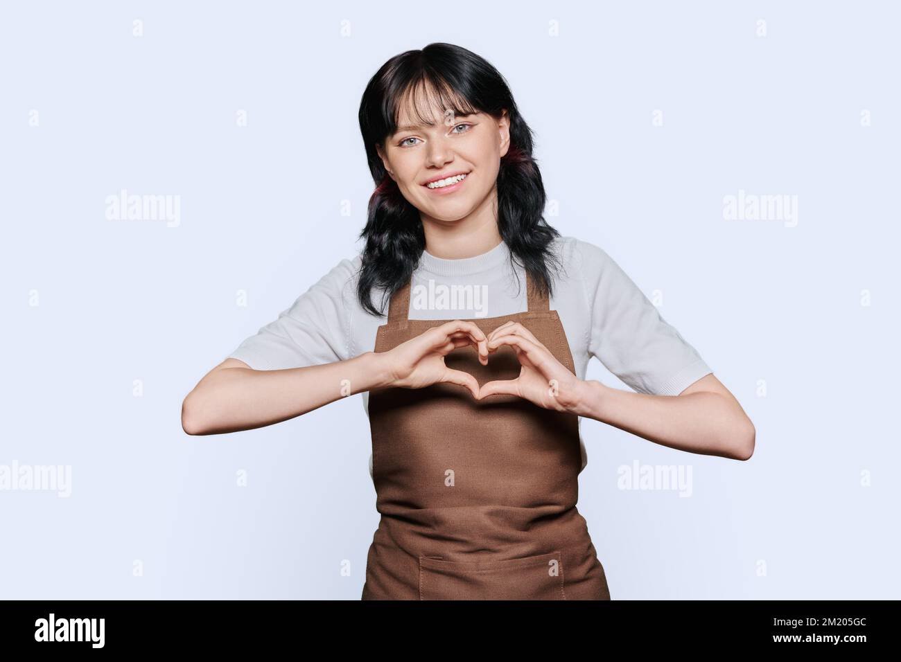 Smiling young female staff in apron showing heart gesture, on white ...