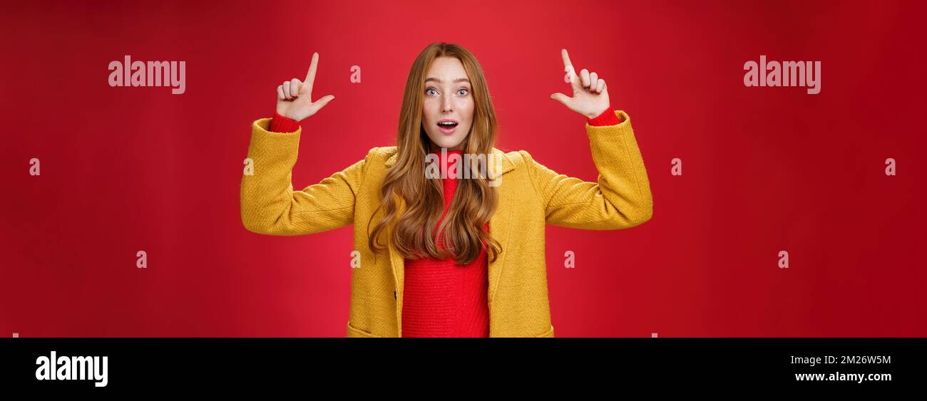 Surprised redhead girl in yellow coat open mouth impressed and thrilled as raising hands and ...