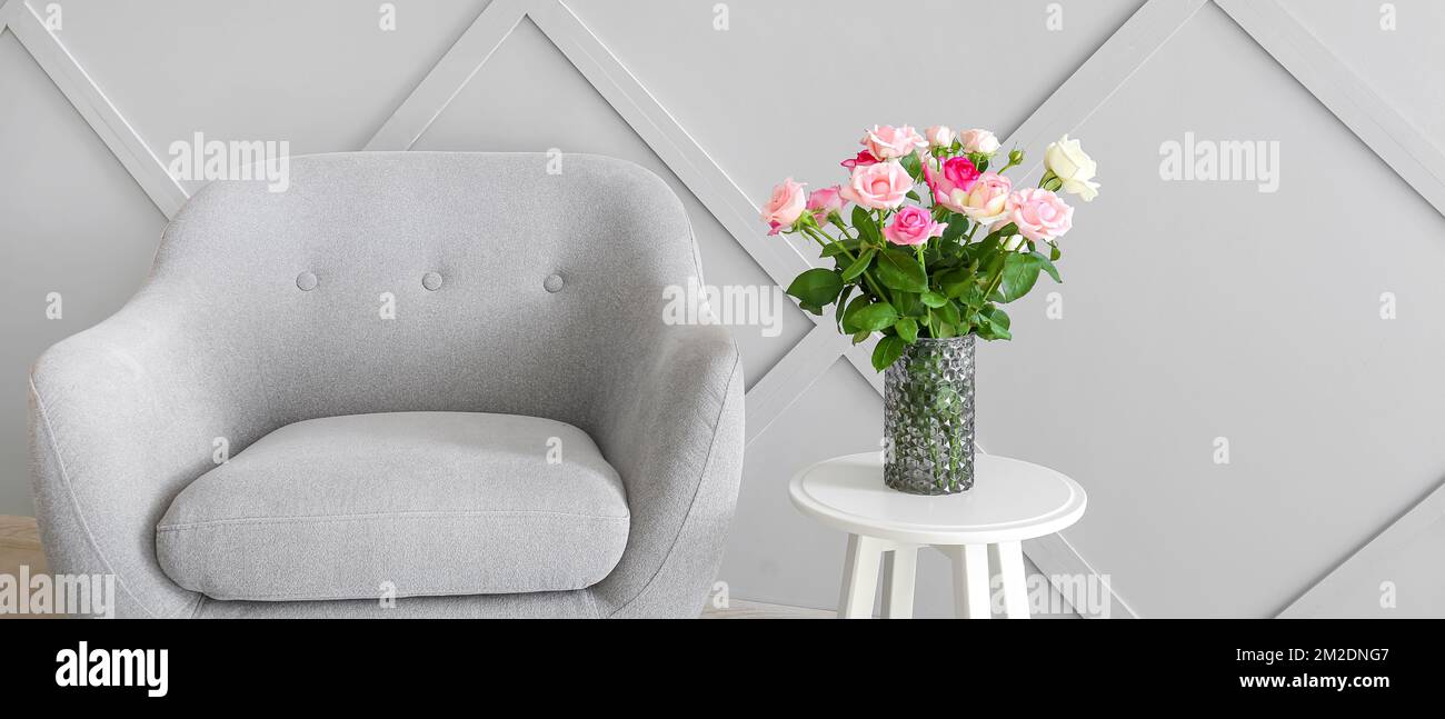 Beautiful rose flowers in vase on table and armchair near grey wall in ...