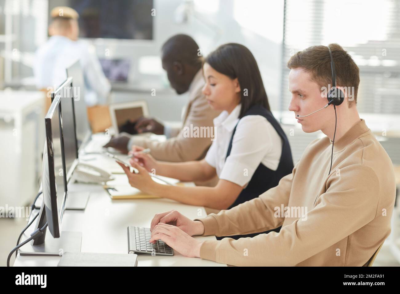 Side view of young people wearing headsets sitting in row while working ...