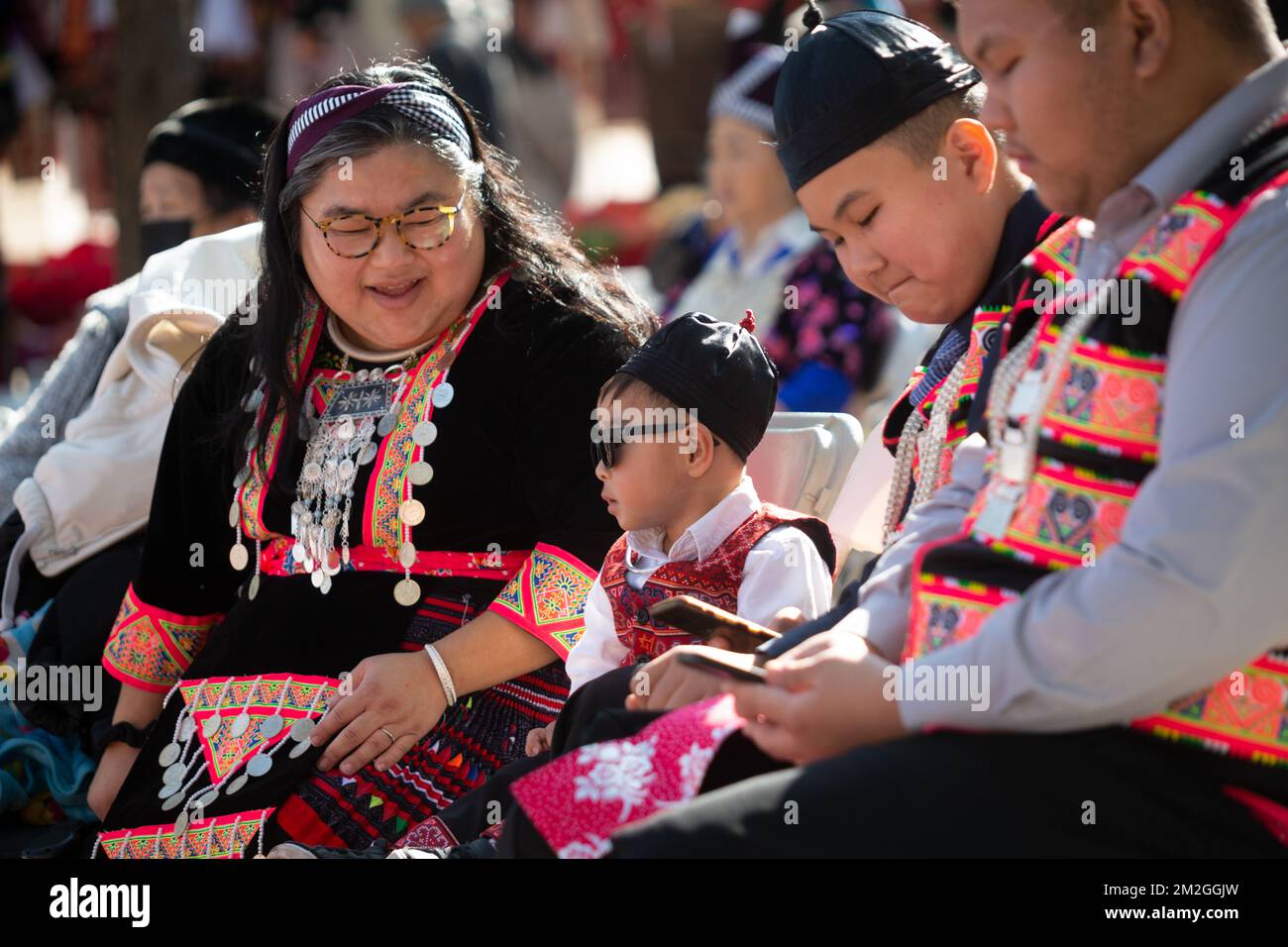 Young boy in sunglasses sitting with his family all dressed in traditional Hmong attire at the ...