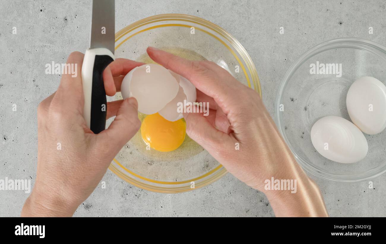 The woman breaks the egg into a glass bowl, a close-up view from above ...