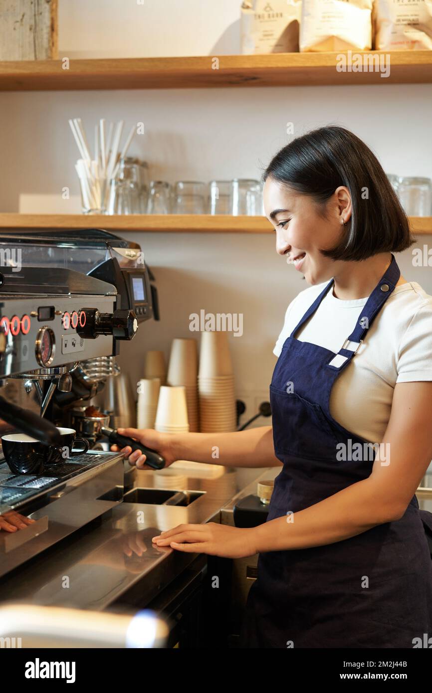 Happy beautiful asian girl, barista in apron works with coffee machine ...