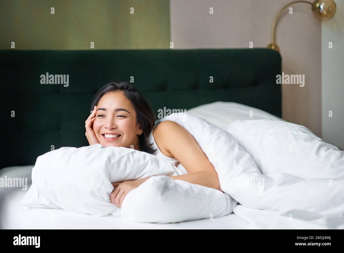 Happy mornings. Beautiful korean woman lying in bed, smiling, waking up ...
