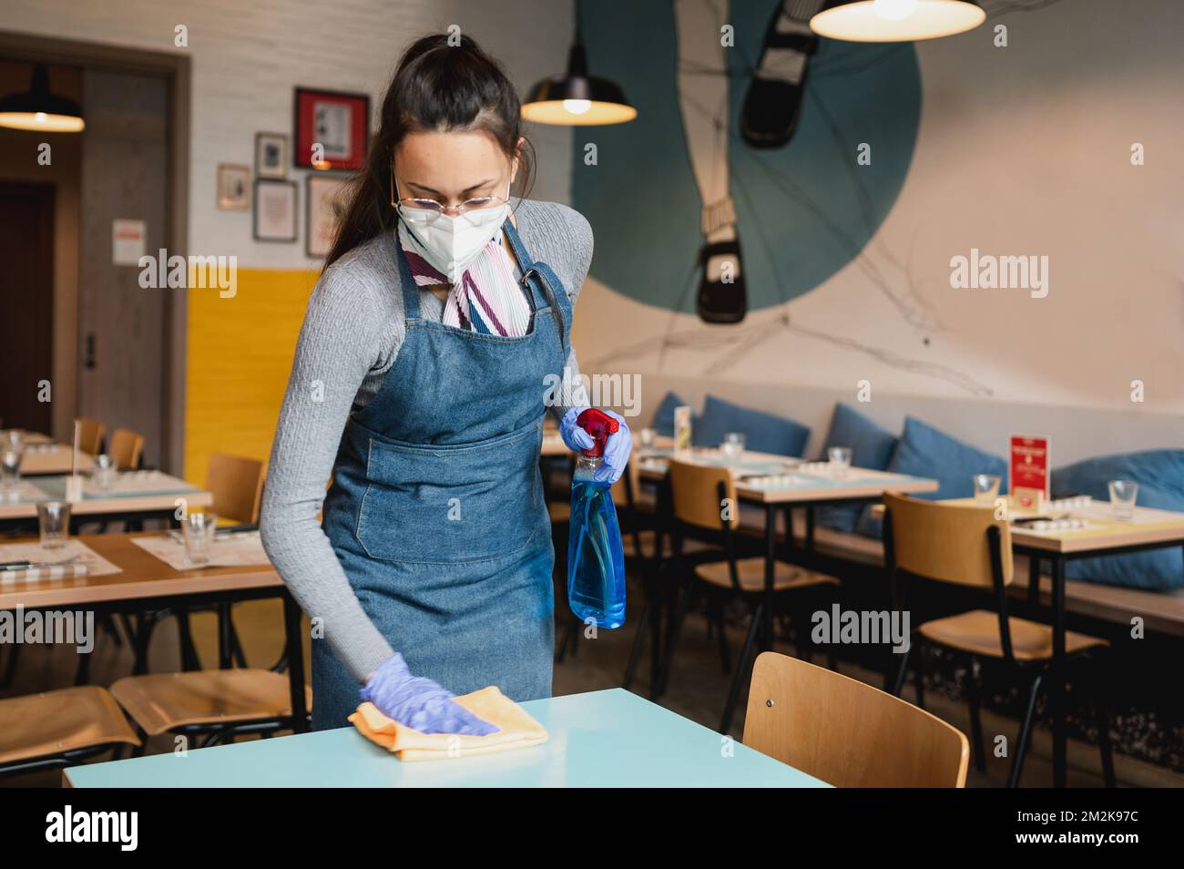 young waitress with apron, face mask and gloves cleaning tables restaurant with sanitizer and ...