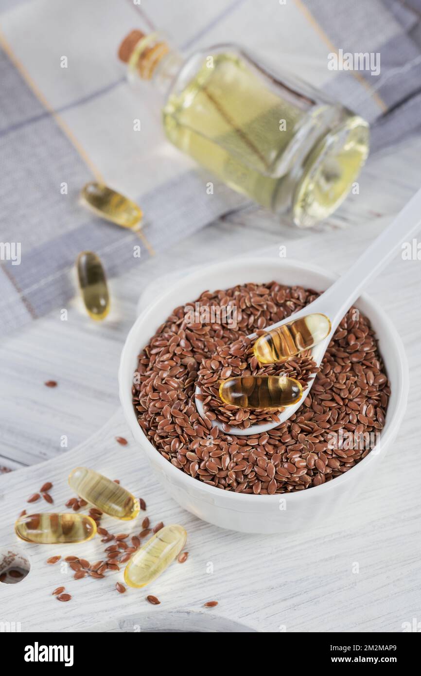 Brown flax seed or linseed in small bowl and gelatin capsules with omega oil on a white wooden ...