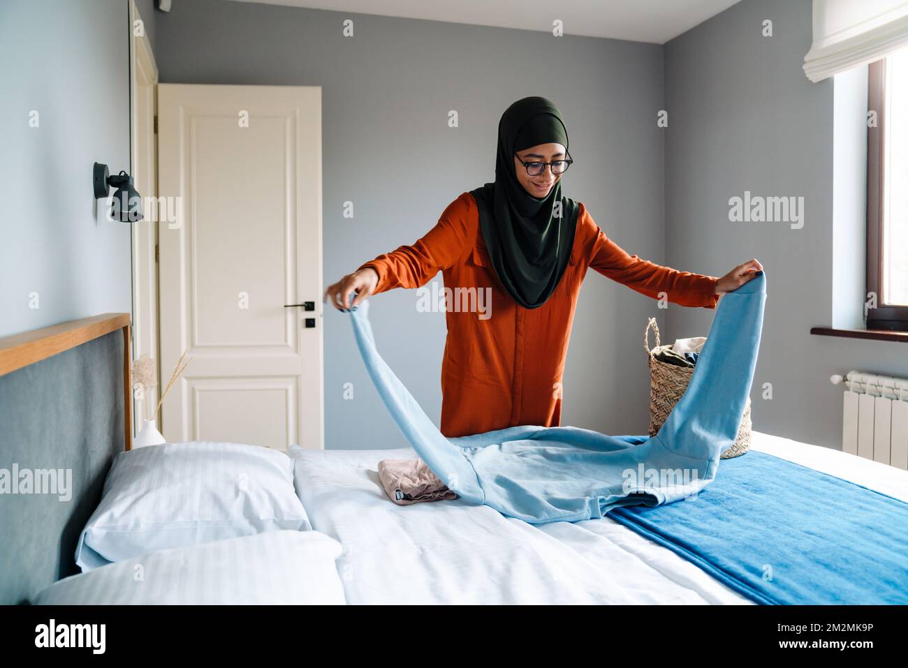 Young beautiful calm woman in hijab and glasses folding clothes at home ...