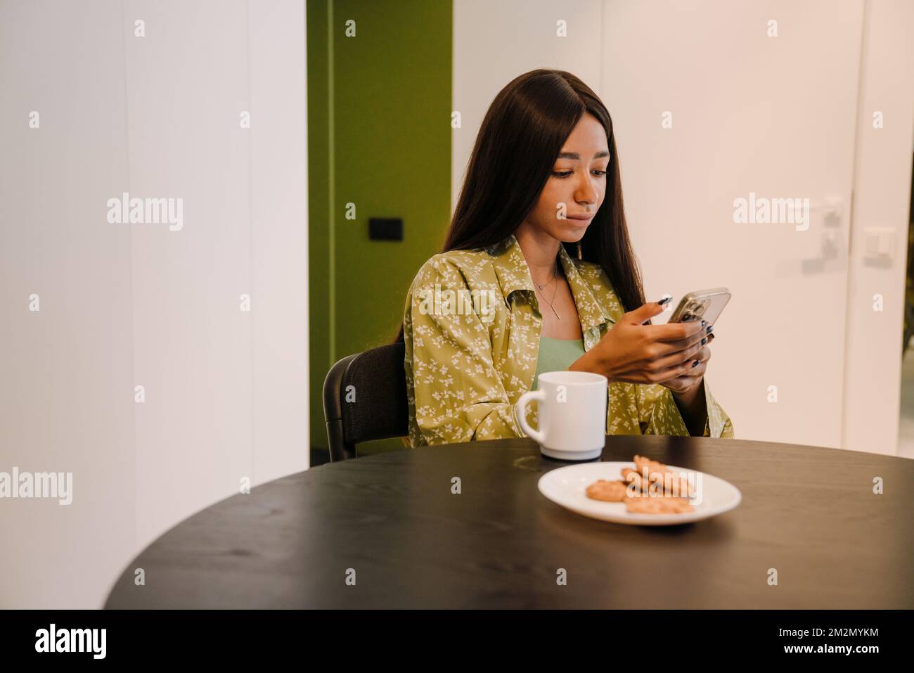 Young beautiful long-haired woman sitting at kitchen table with cup of ...