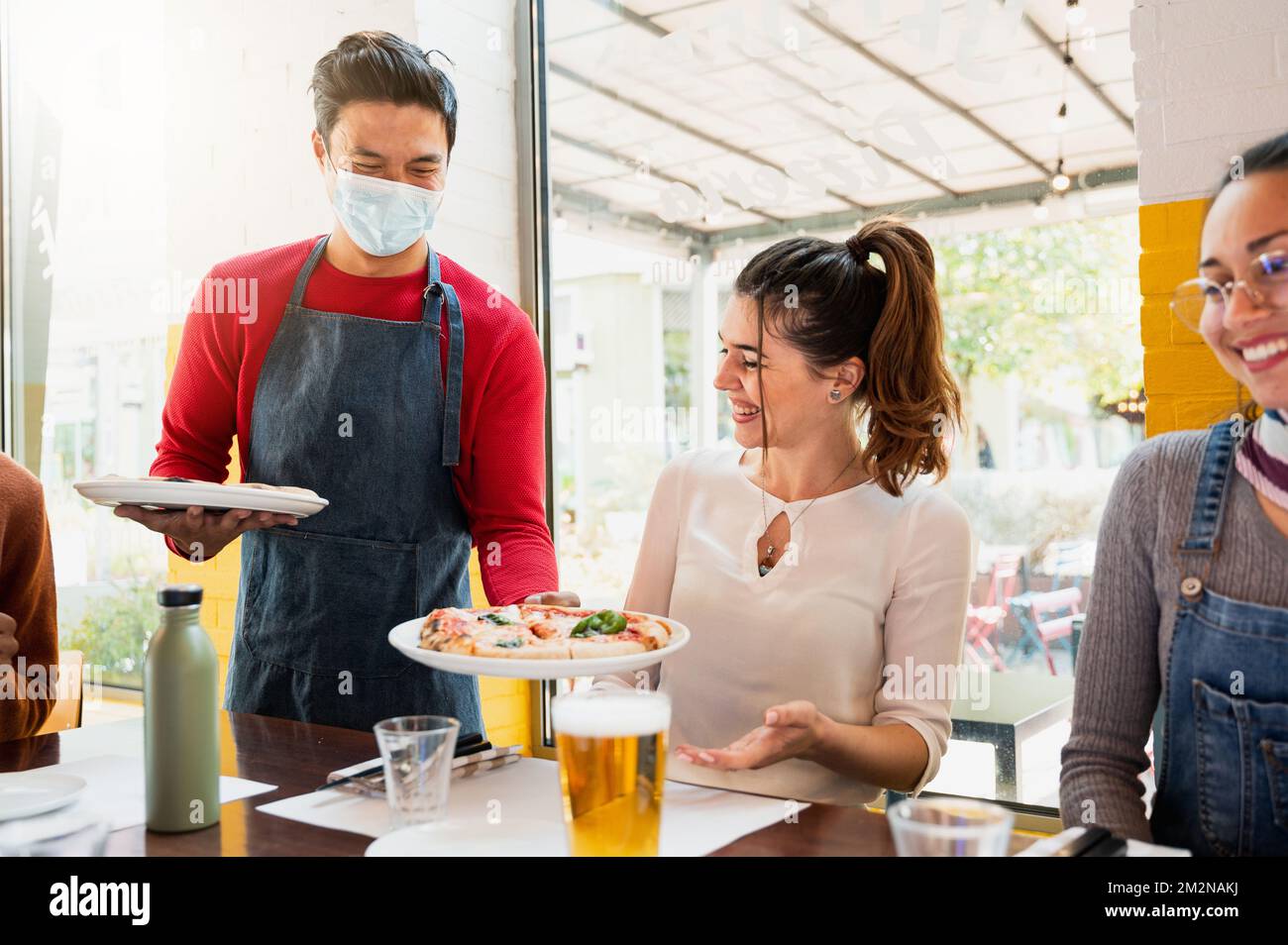 Waiter with protective face mask serving pizza to smiling females in a ...