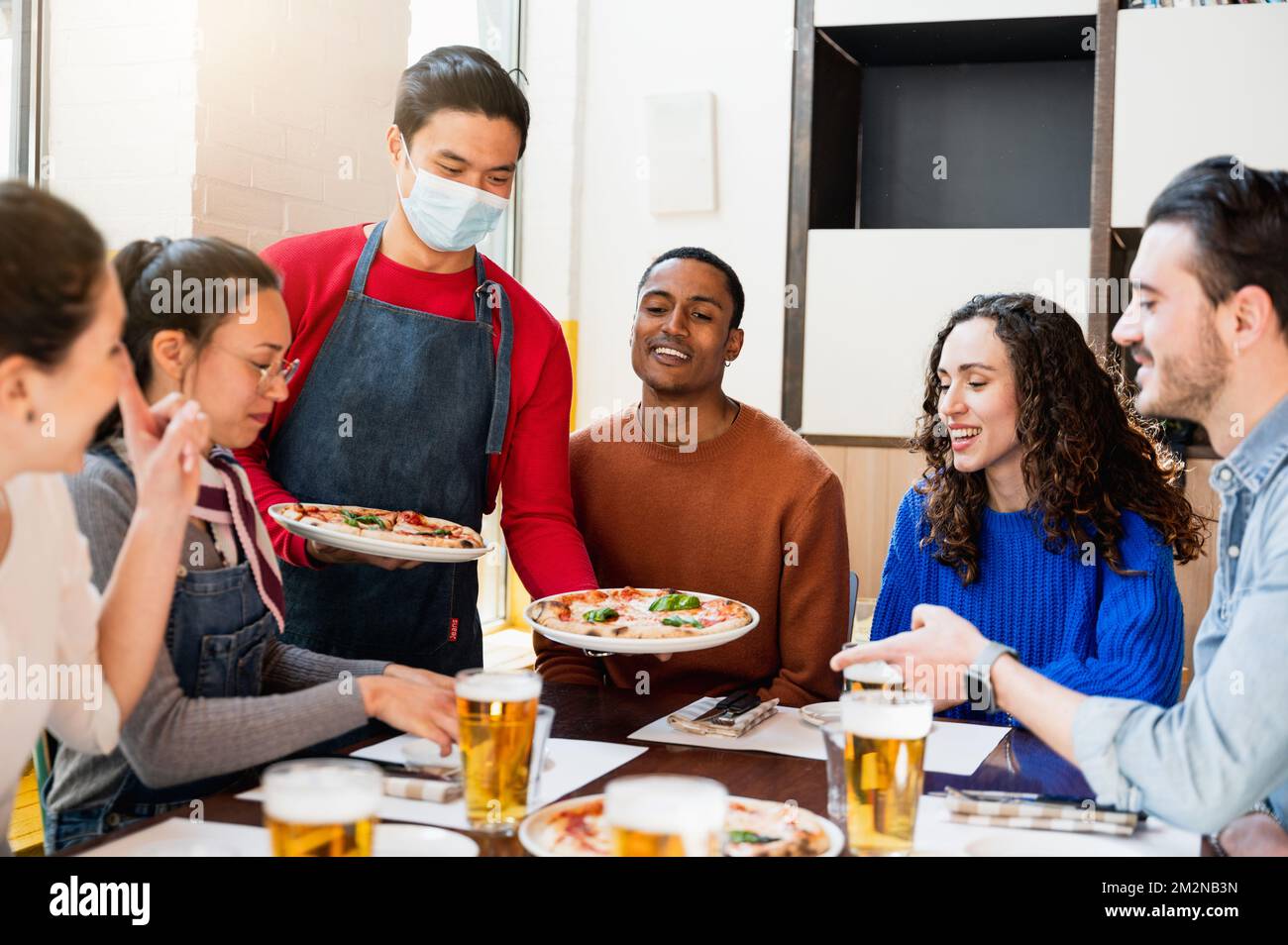 Asian young waiter wearing protective face mask serving pizza to a ...
