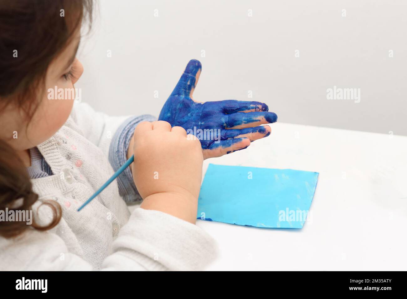 Child paints her own hand with blue paint and paintbrush getting messy ...