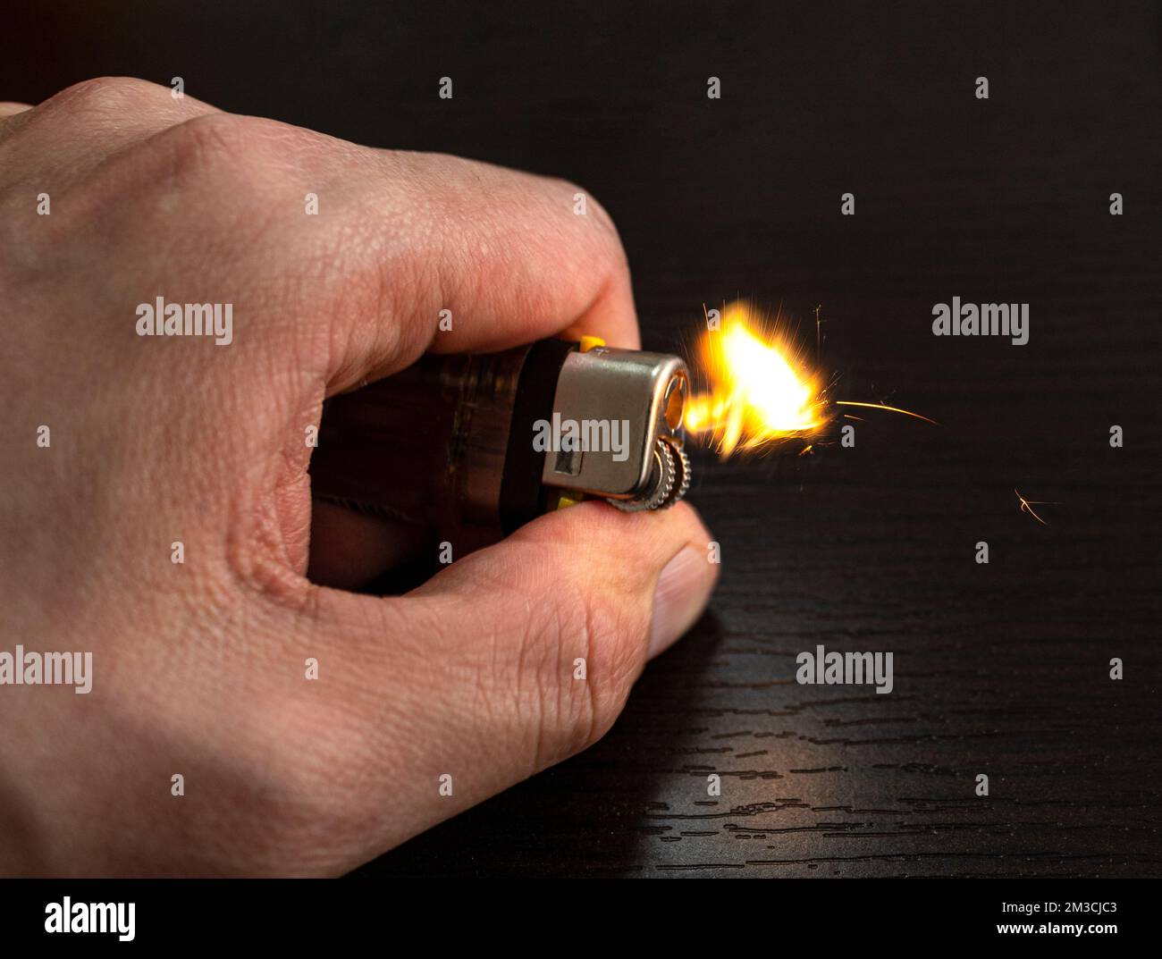 Closeup to a male hand holding a yellow lighter with flame over a dark ...