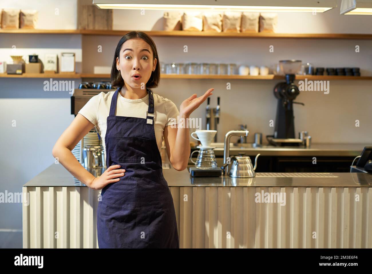 Image of excited asian female barista, looking surprised, pointing at ...