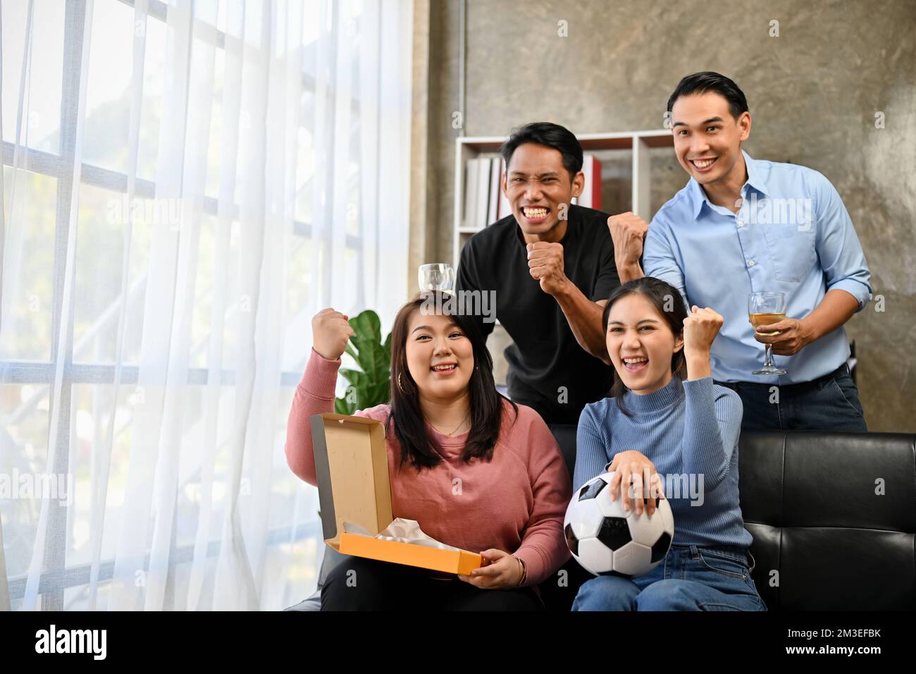 Group of joyful Asian friends sit on sofa watching and cheering an ...