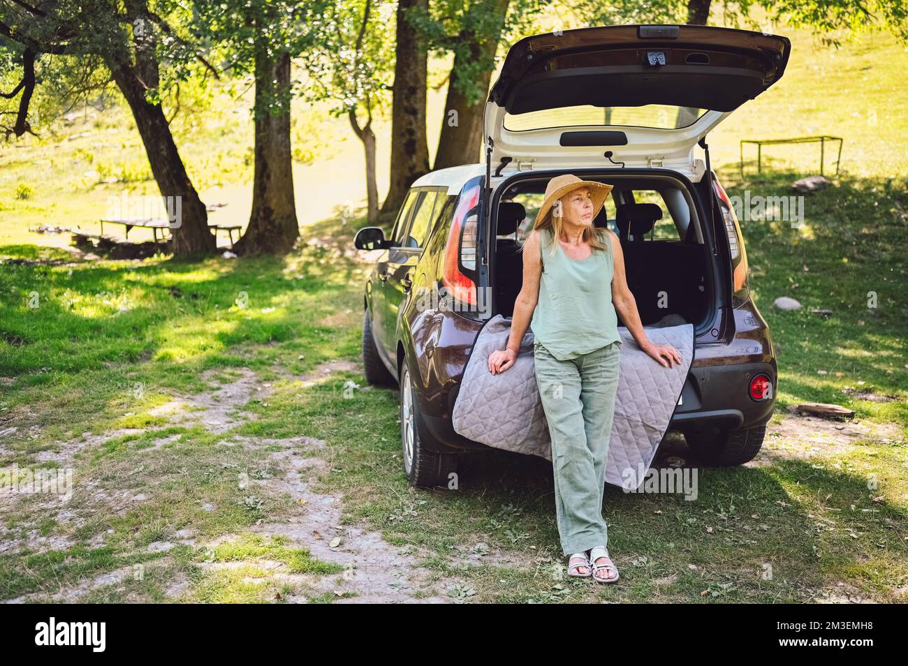Happy senior woman in straw hat sitting in car trunk or boot outdoors ...