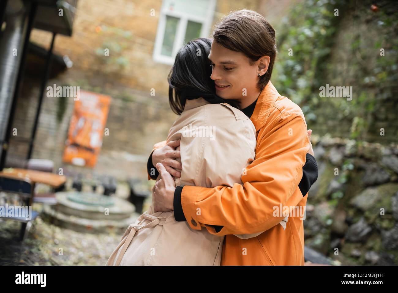 Smiling man hugging girlfriend in trench coat on terrace of cafe,stock ...