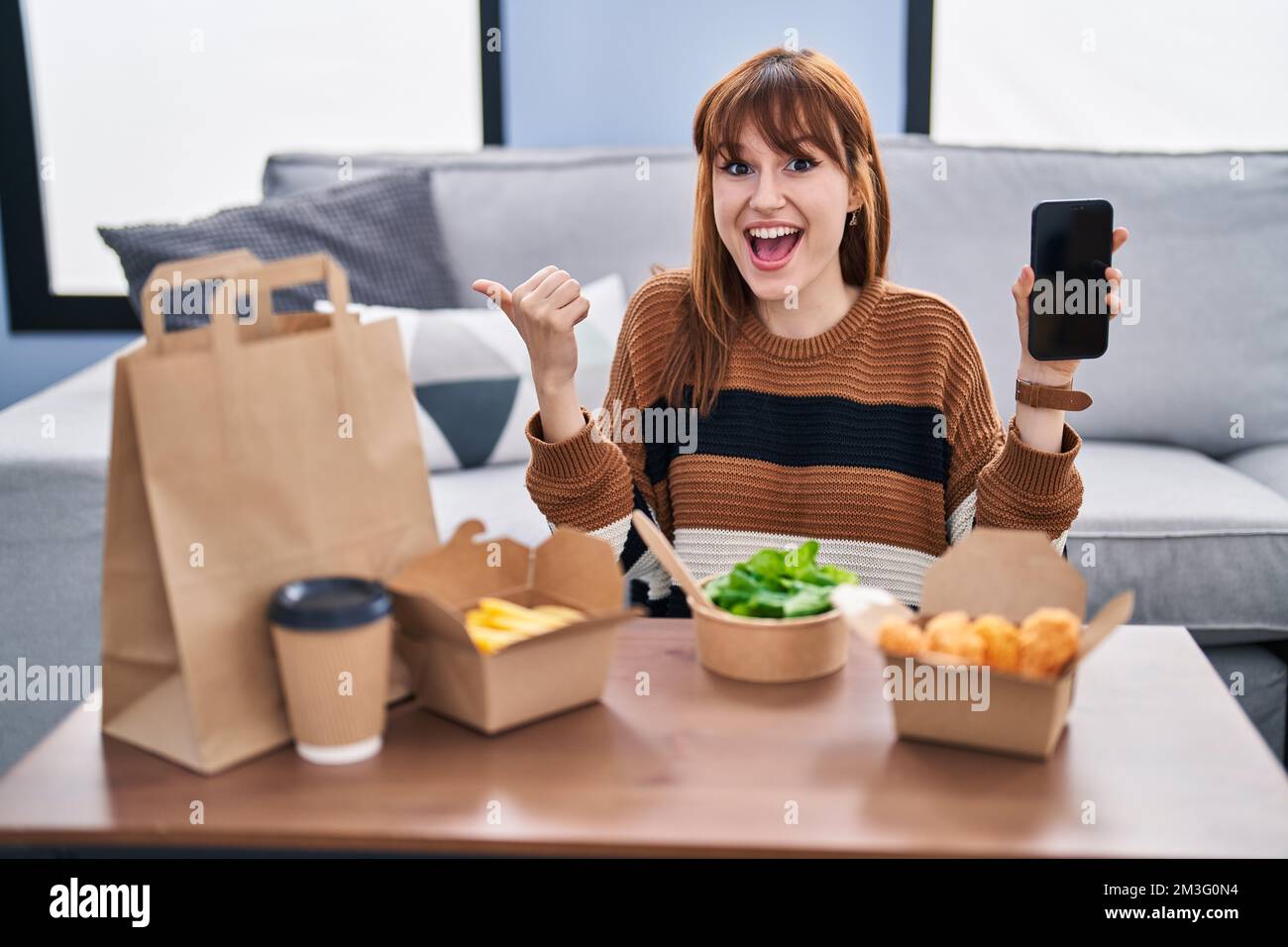 Young beautiful woman eating delivery food at the living room pointing ...