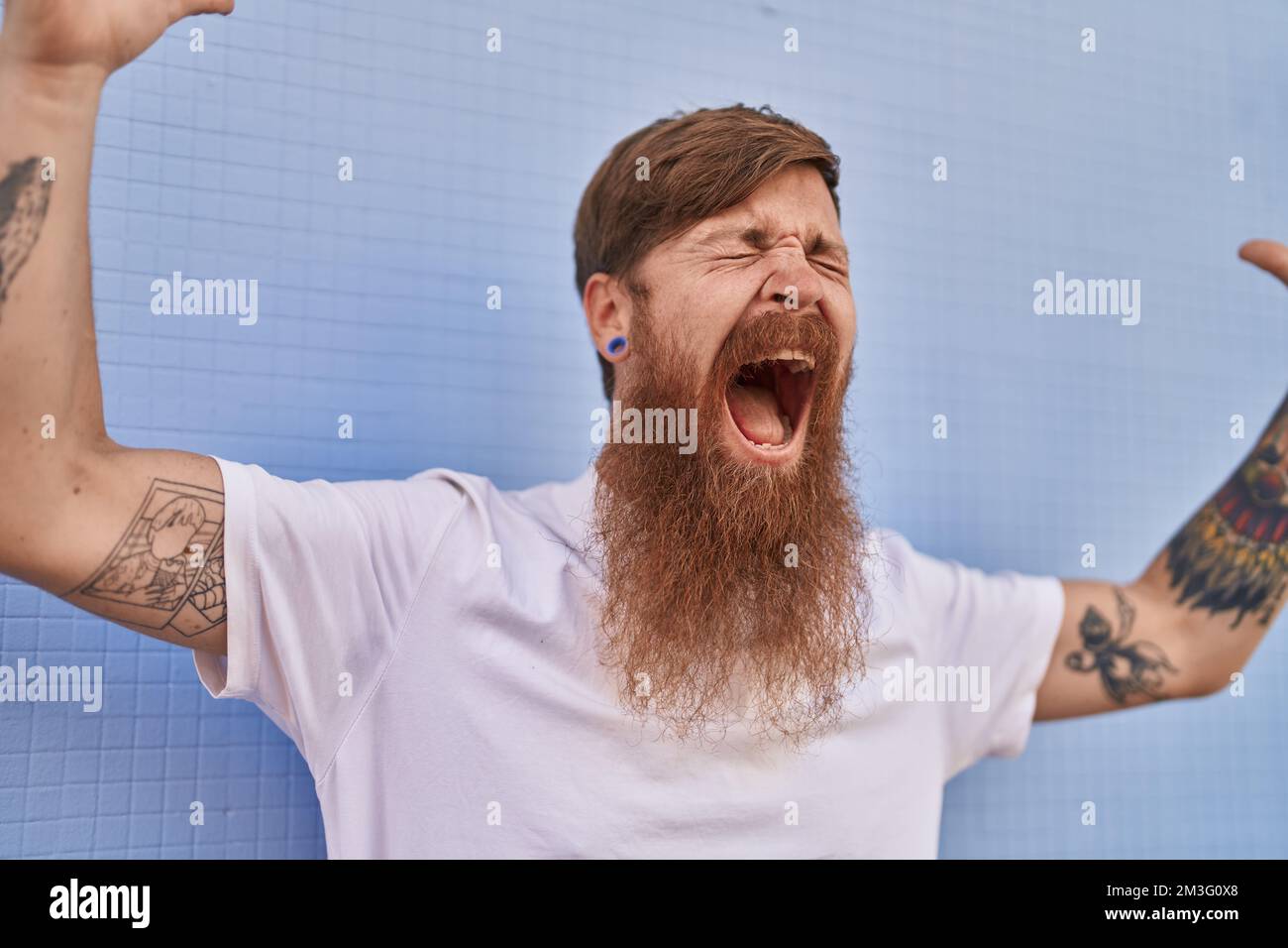 Young redhead man looking to the side shouting over isolated blue ...