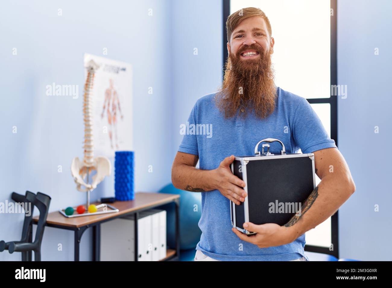 Young redhead man wearing physiotherapist uniform holding briefcase at ...