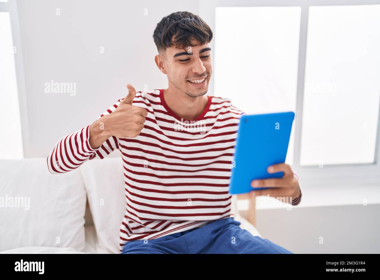 Young hispanic man using touchpad sitting on the bed smiling happy and positive, thumb up doing ...