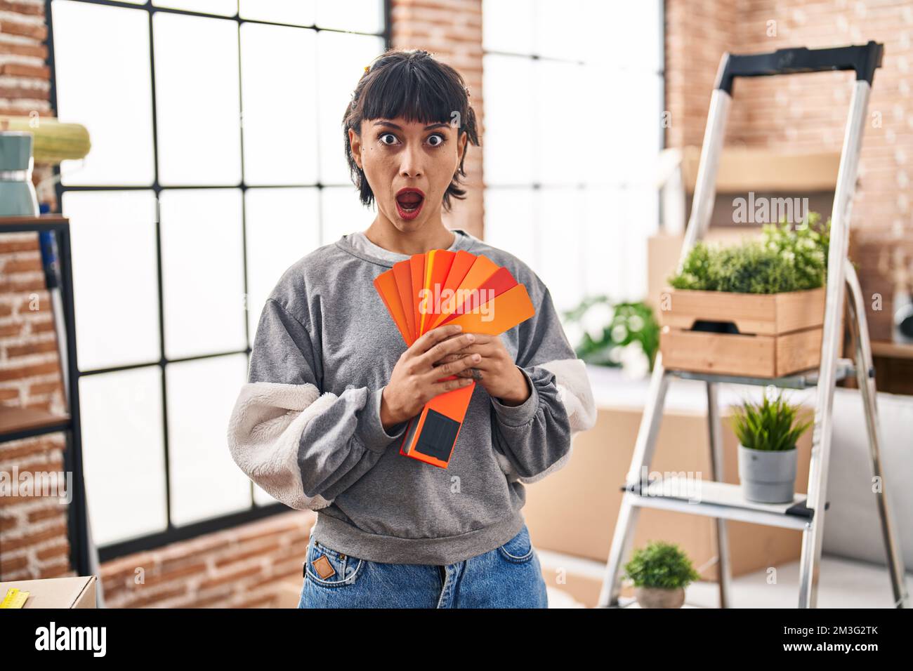 Young hispanic woman moving to a new home choosing paint in shock face ...
