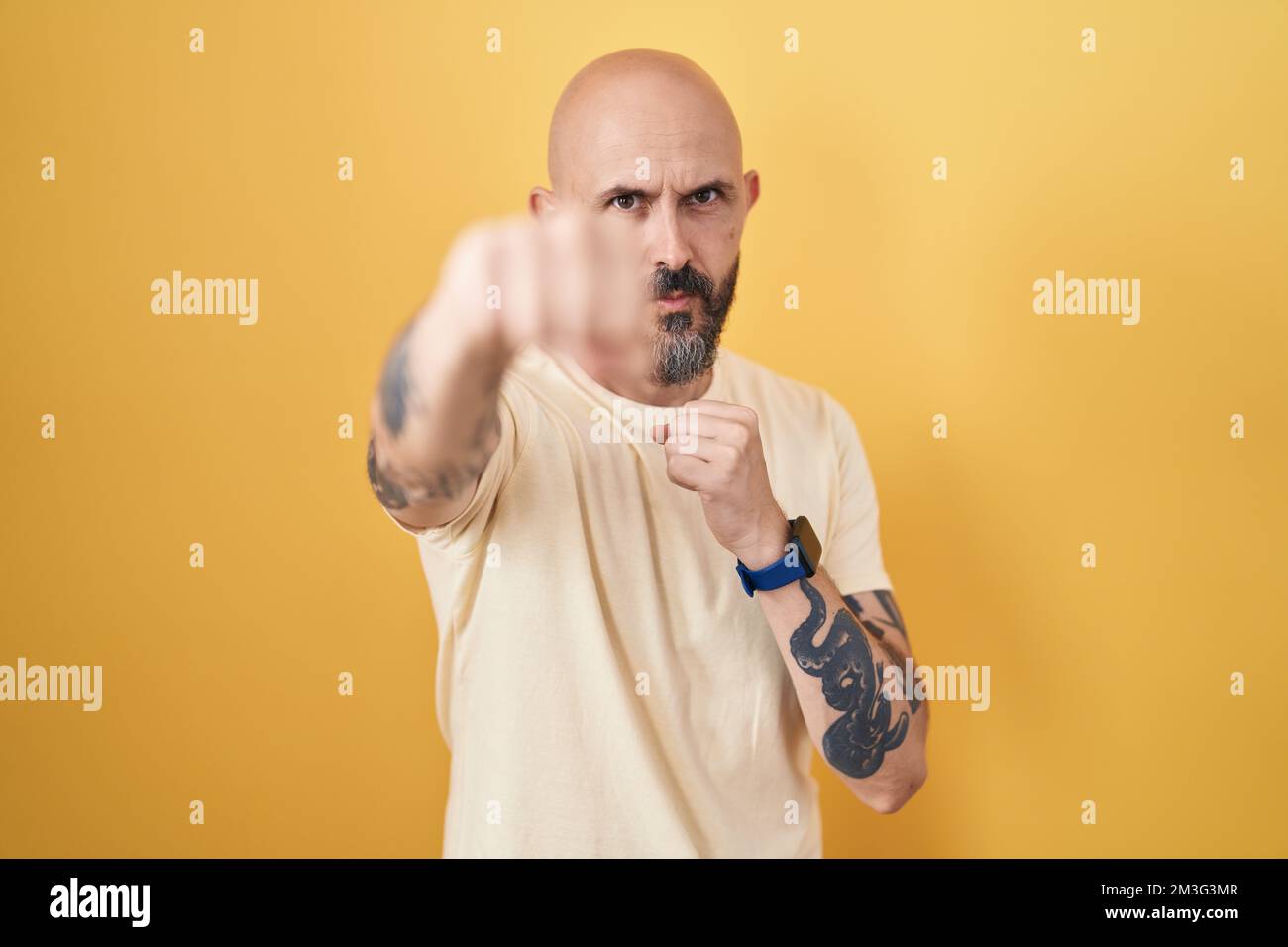 Hispanic man with tattoos standing over yellow background punching fist ...