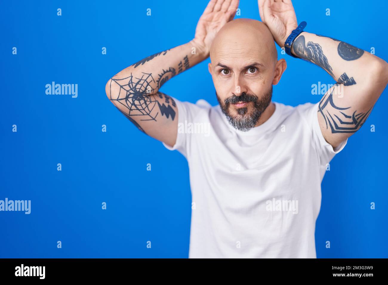 Hispanic man with tattoos standing over blue background doing bunny ...