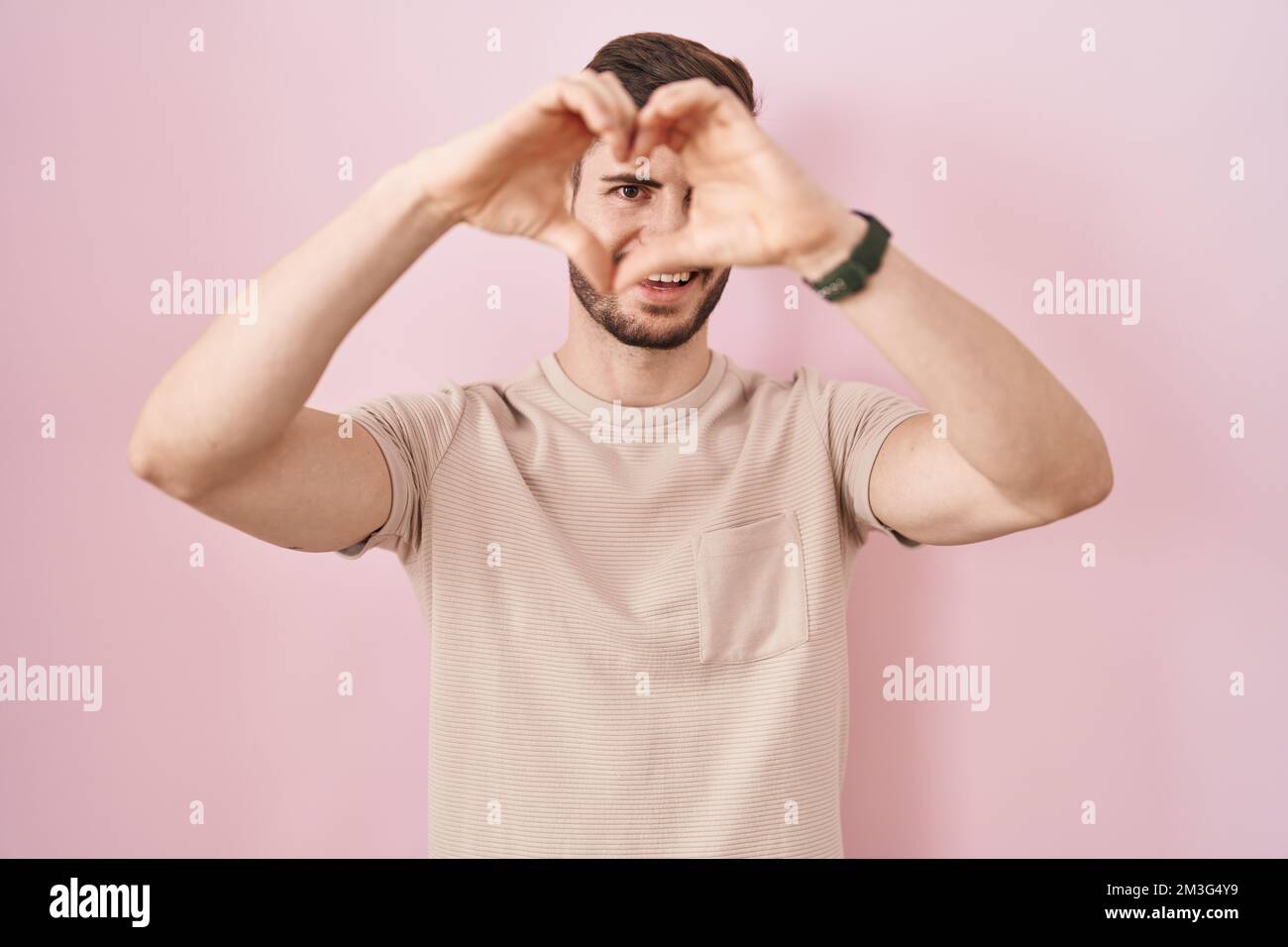 Hispanic man with beard standing over pink background doing heart shape ...